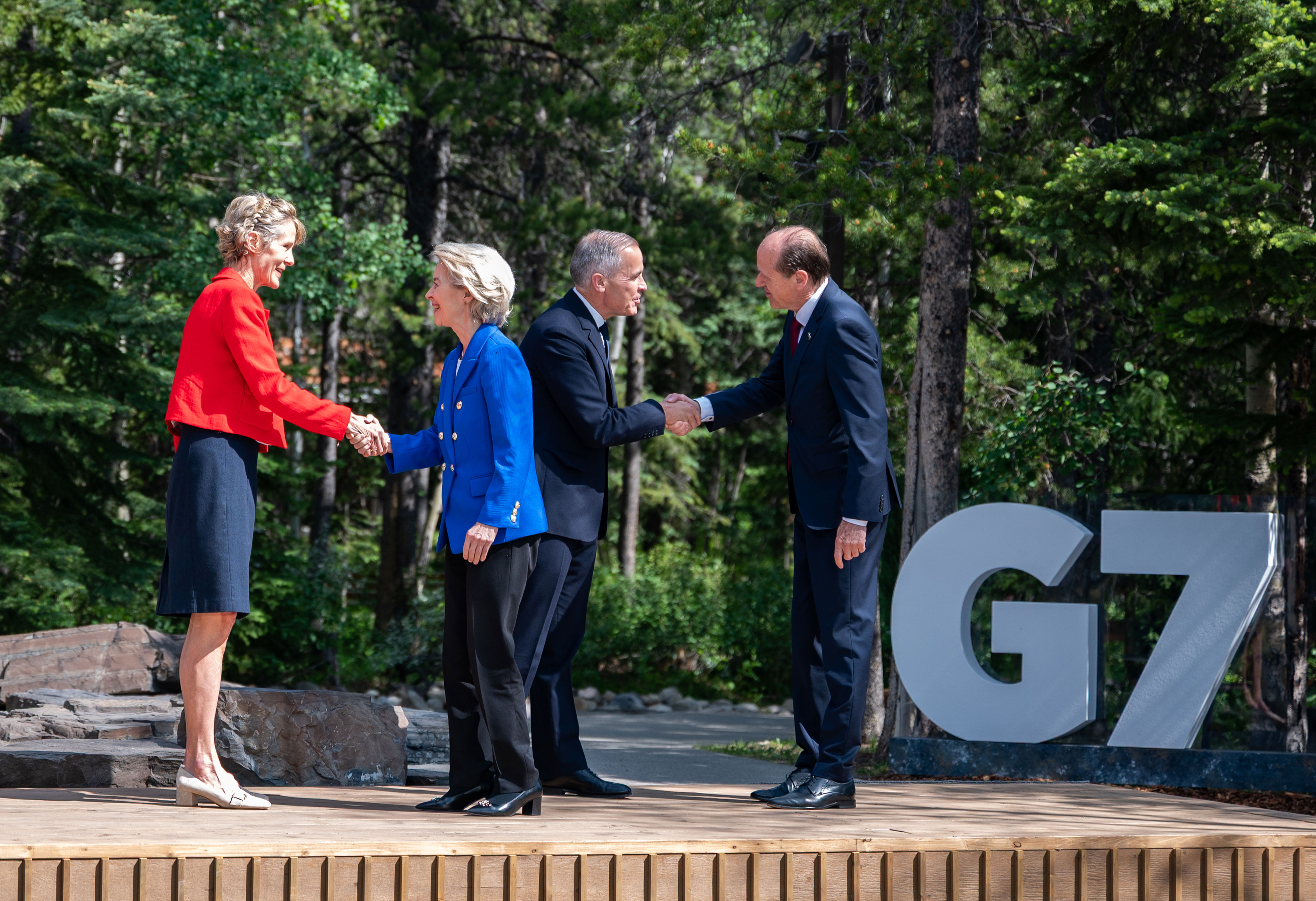 People shaking hands on a stage in a wooded area, with a large 3D white ...